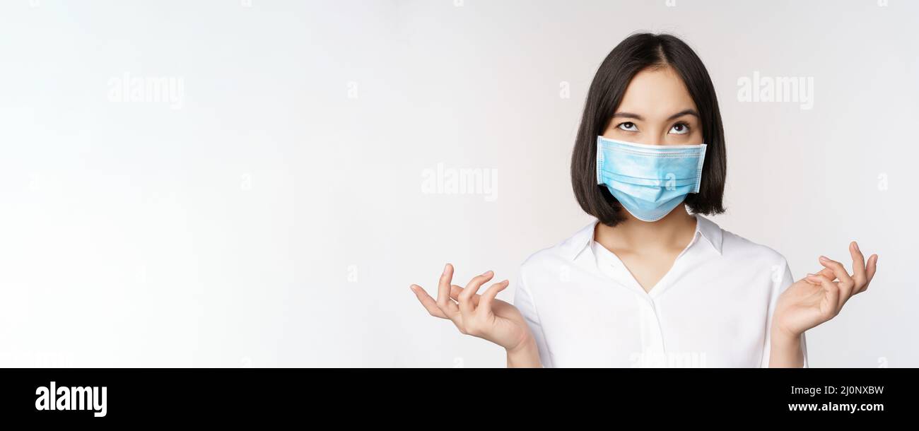 Annoyed asian woman in medical face mask, shrugging and looking up with ...