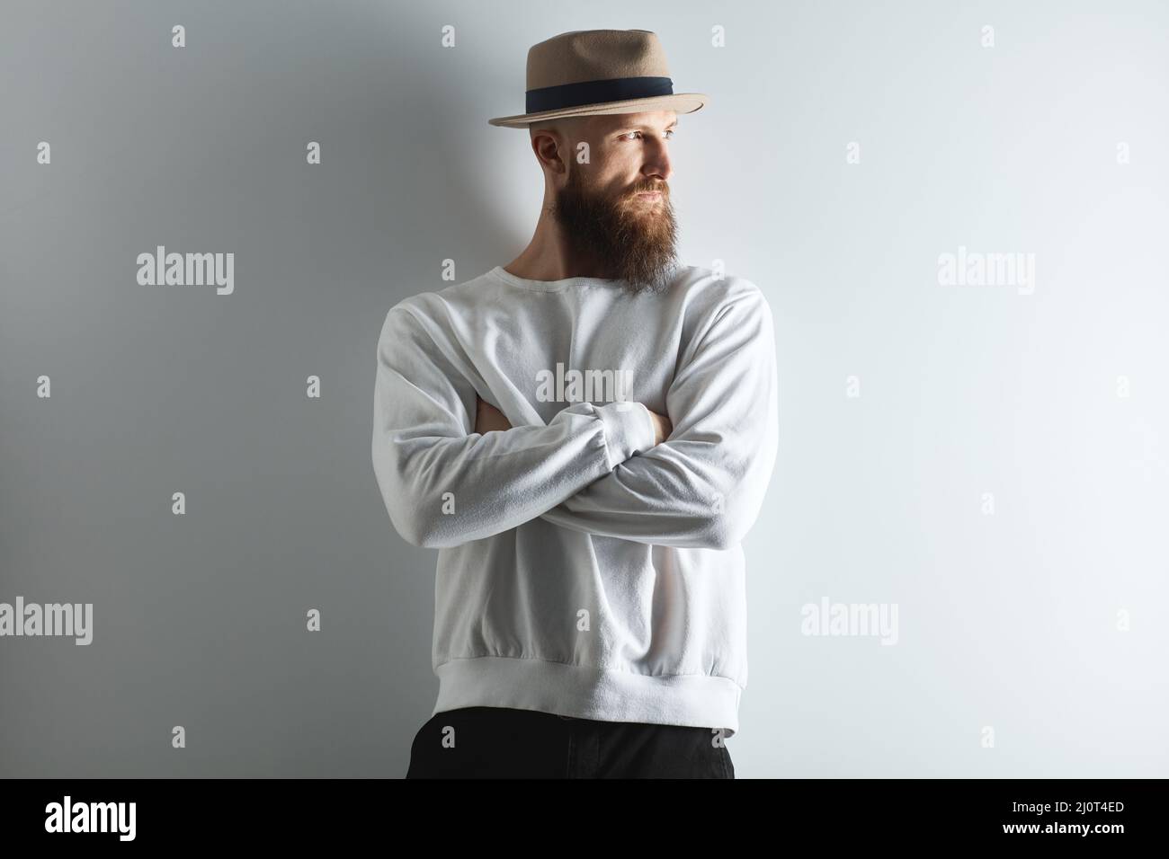 Serious bearded man in hat standing against white wall arms crossed ...
