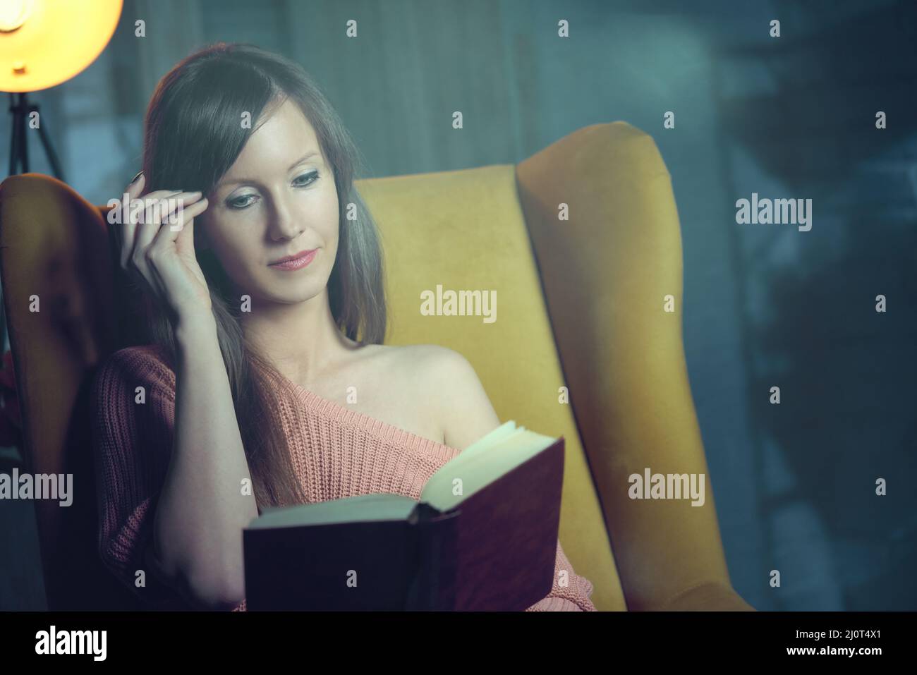 White woman reading book at home sitting in armchair in cosy room ...