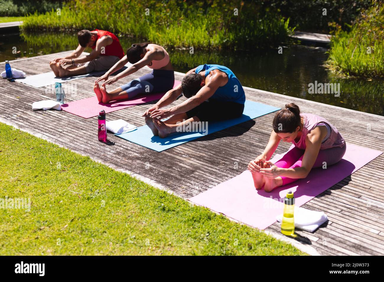 Men and women touching toes while practicing yoga on floorboard by pond ...