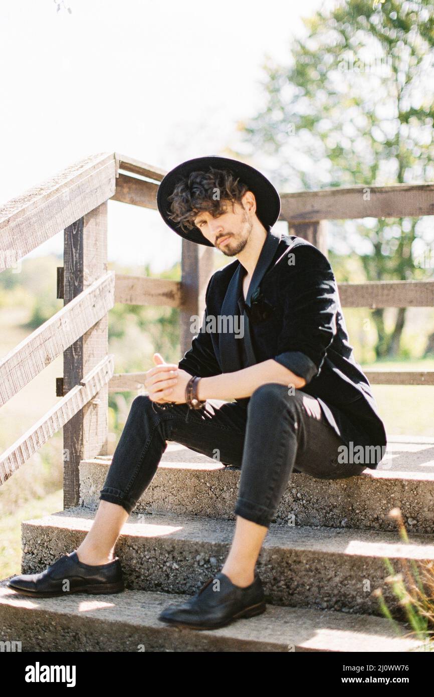 Man in a black jacket and hat sits on the steps near the wooden railing ...