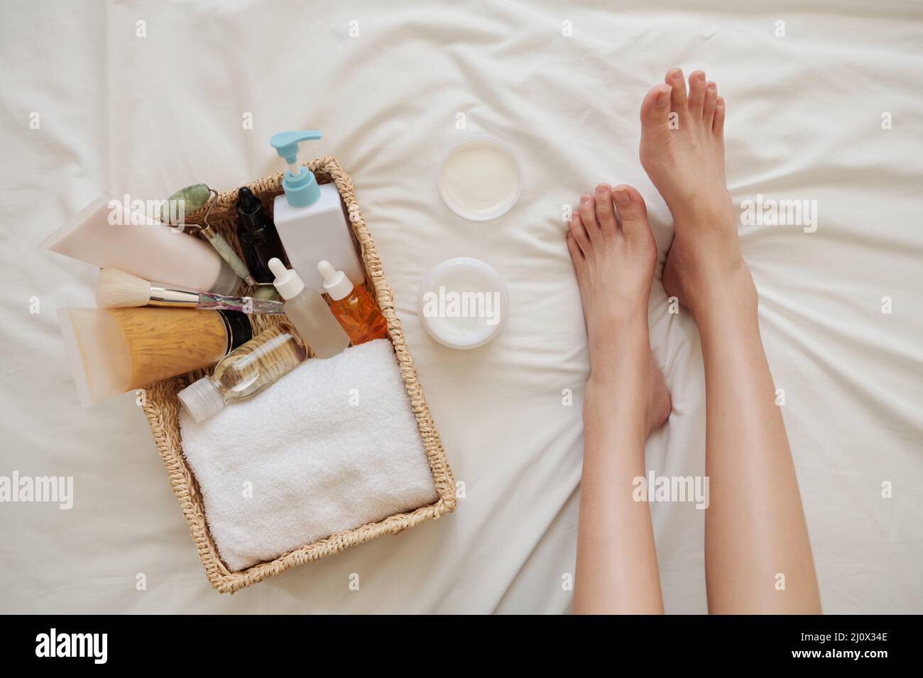 Feet of young woman on bed next to basket of skincare products like ...