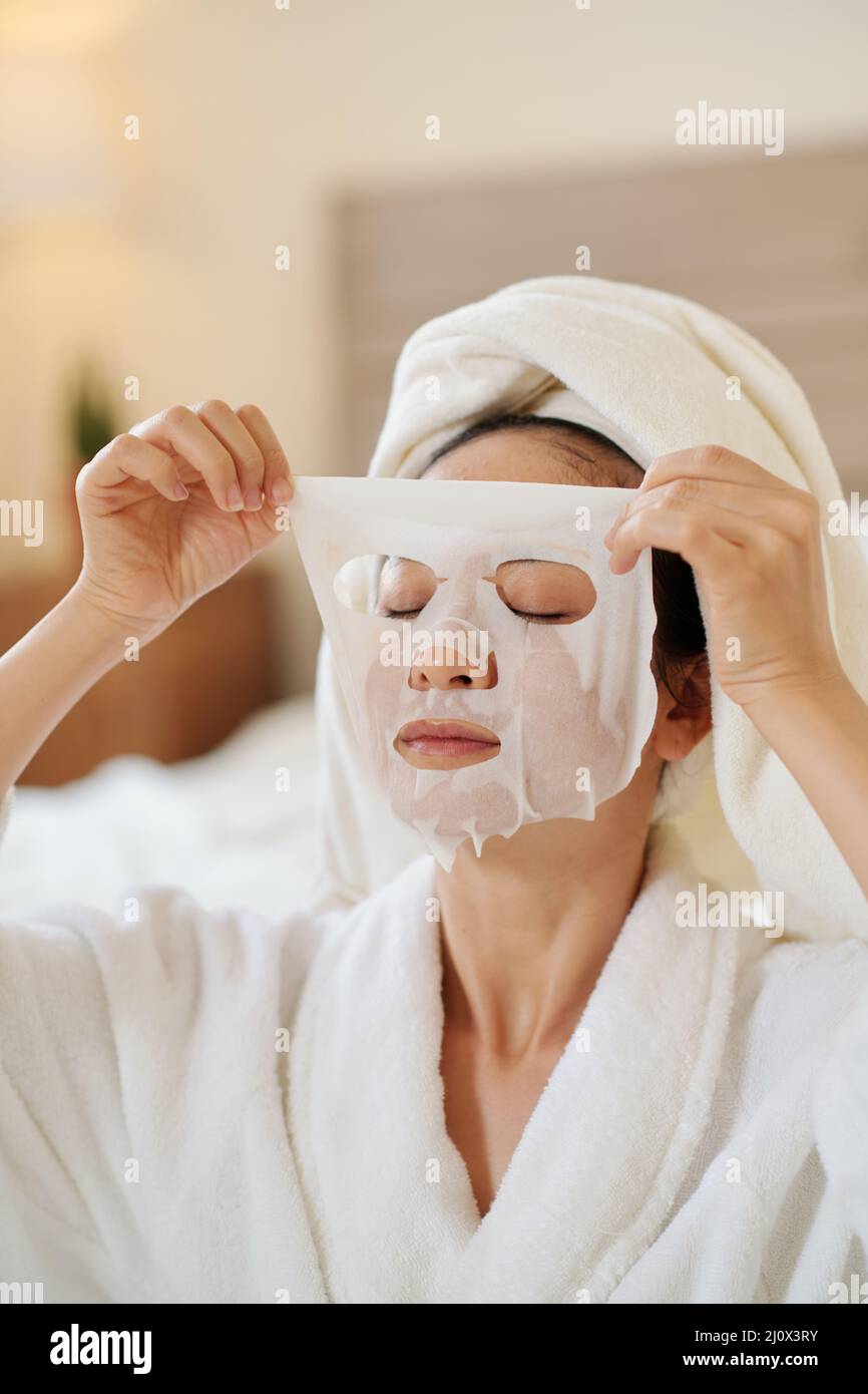 Calm young woman applying cooling sheet mask on face in the morning ...