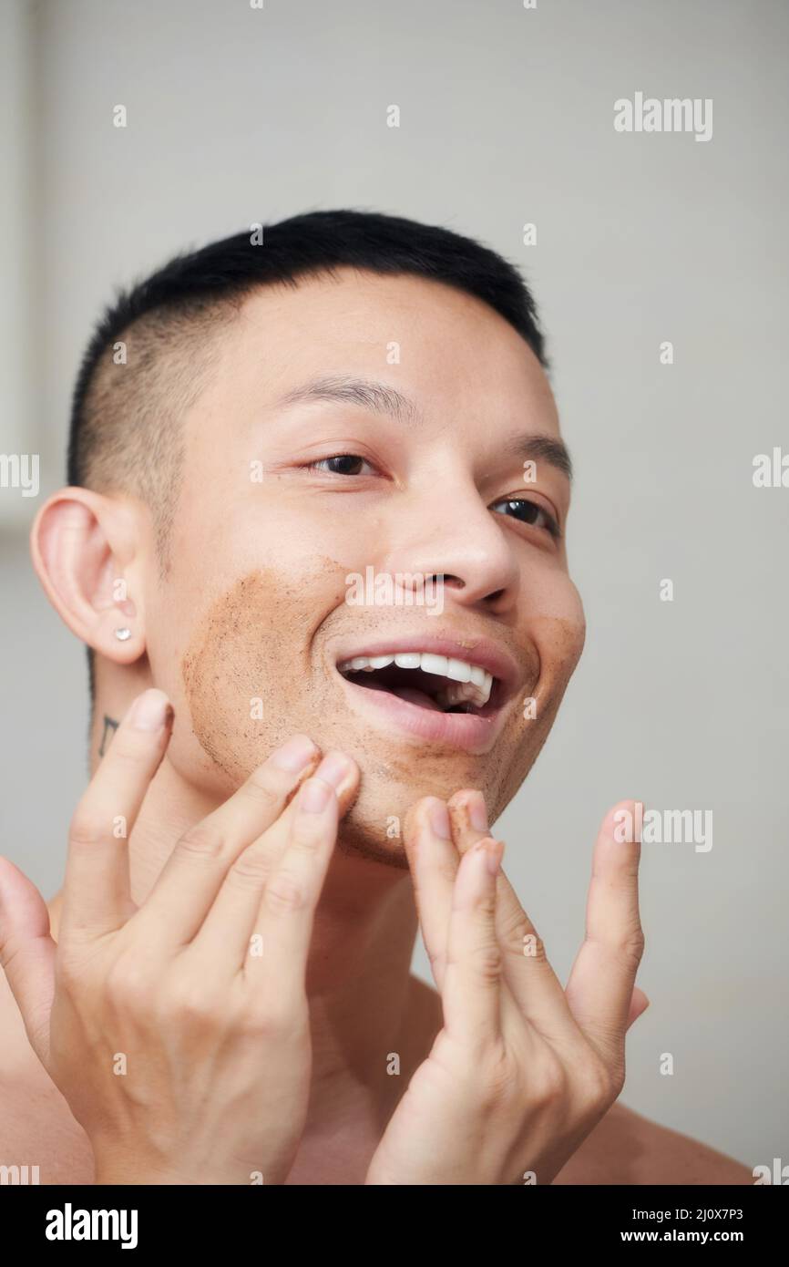 Happly young man applying red clay mask on chin to deep clean pores ...