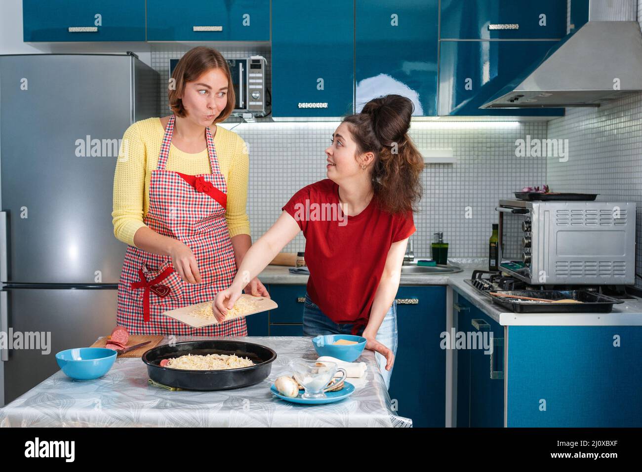 Portrait of two young women cook pizza, sprinkling cheese on top of it ...