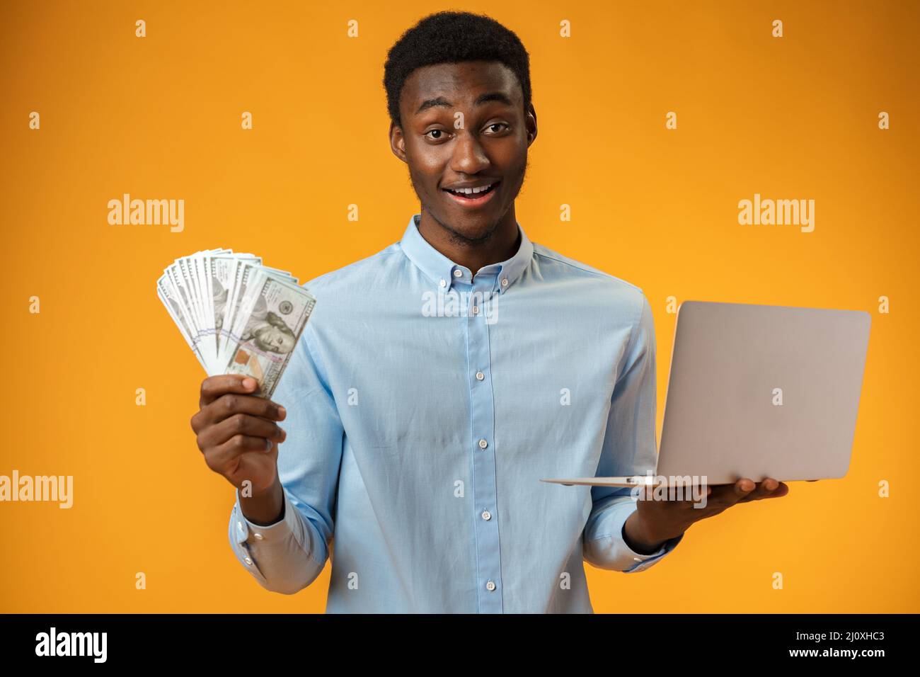 Portrait of joyful black man with laptop and money standing over yellow ...