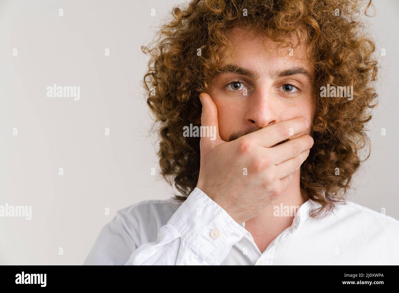 Young curly man with ginger hair posing and covering his mouth isolated ...