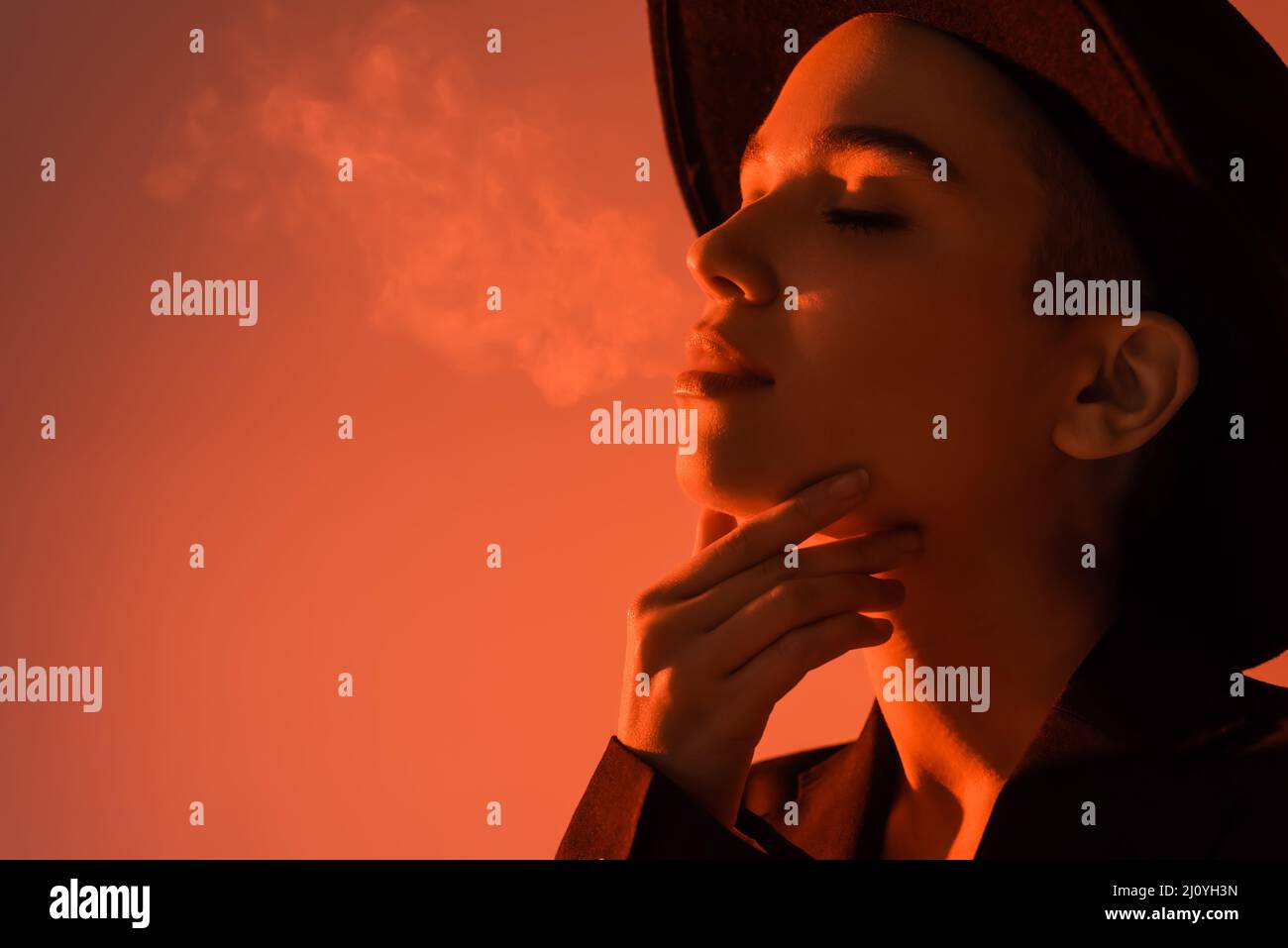 young woman with closed eyes touching chin while exhaling smoke on ...