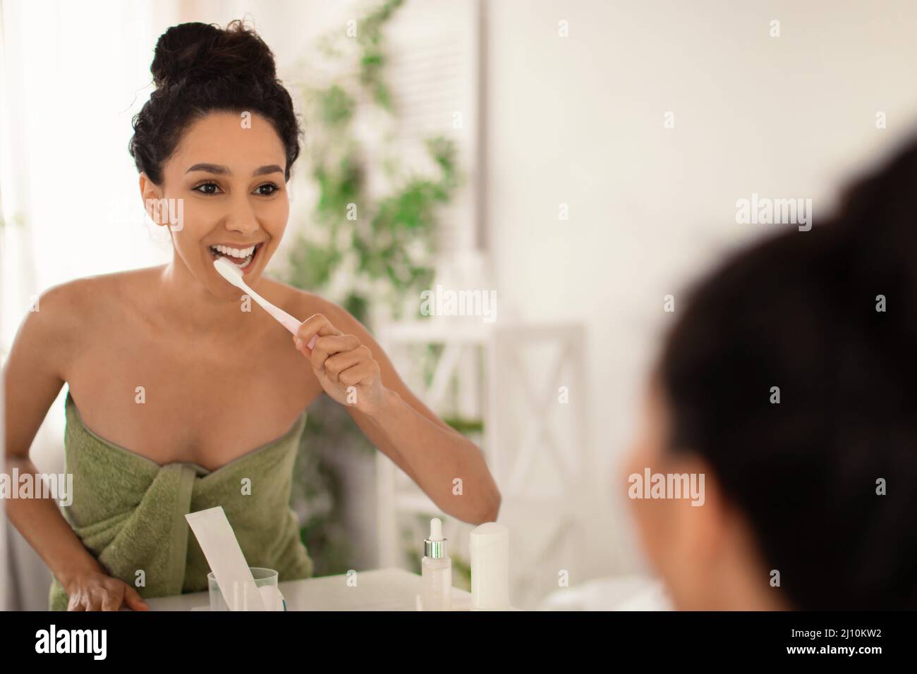 Lovely young lady in towel brushing teeth with toothbrush near mirror ...