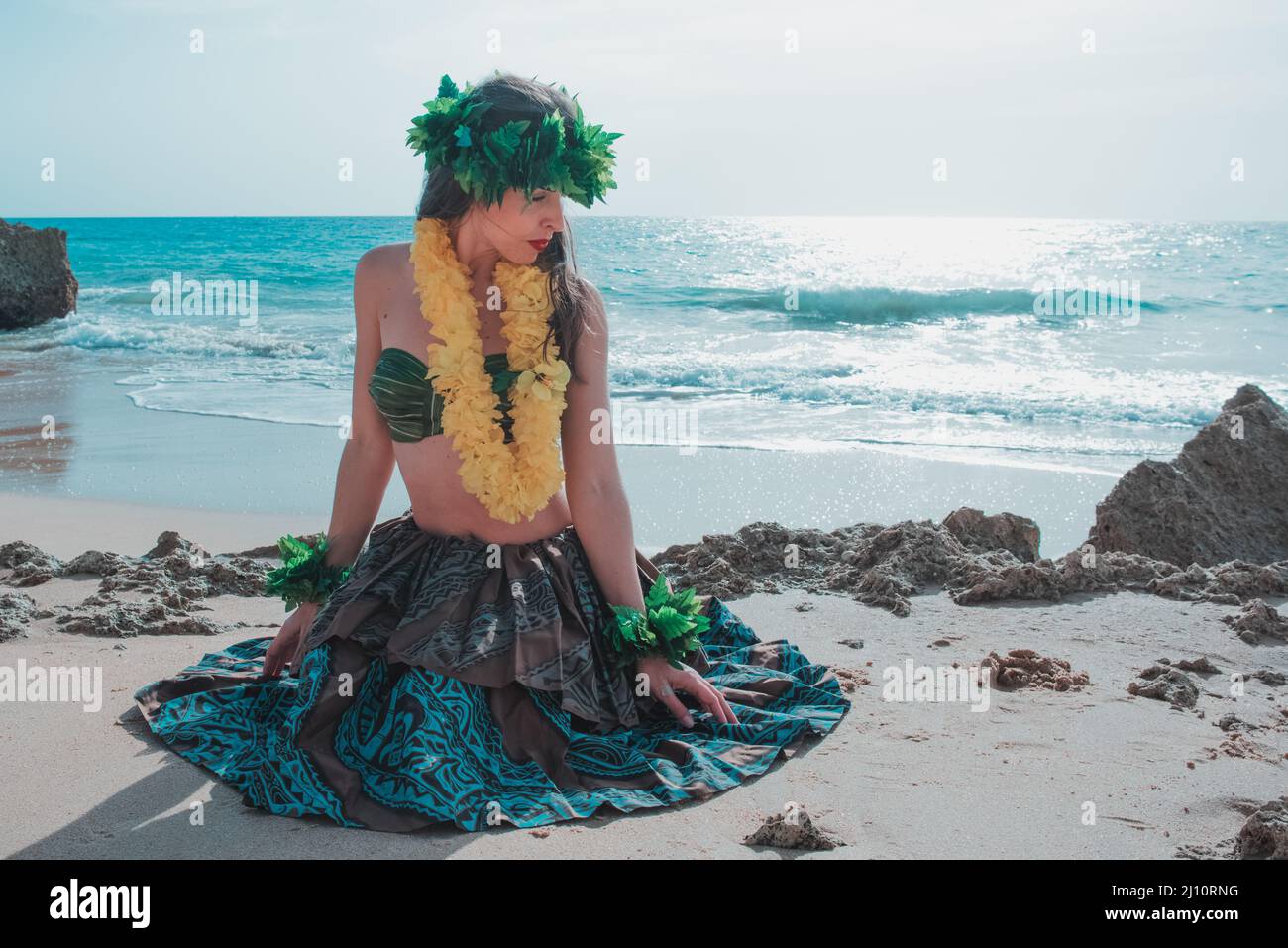 Attractive woman in a Hula costume sitting on the sand at a rocky beach ...