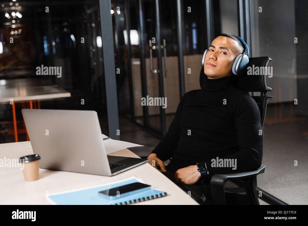 Overtime working. Asian man with closed eyes is resting at work at laptop. Tired male programmer ...