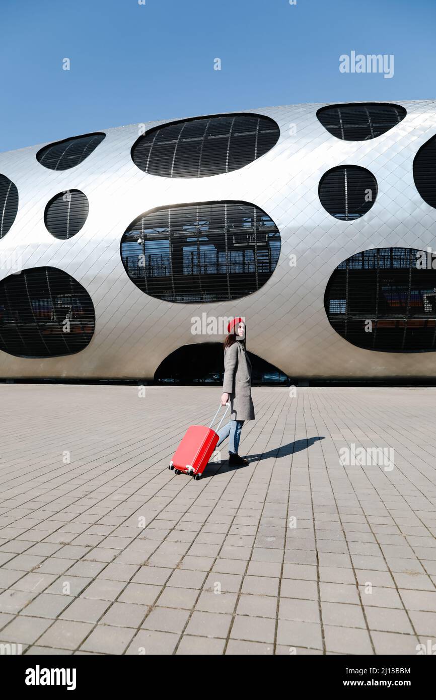 young woman tourist with a red travel suitcase by the futuristic facade ...