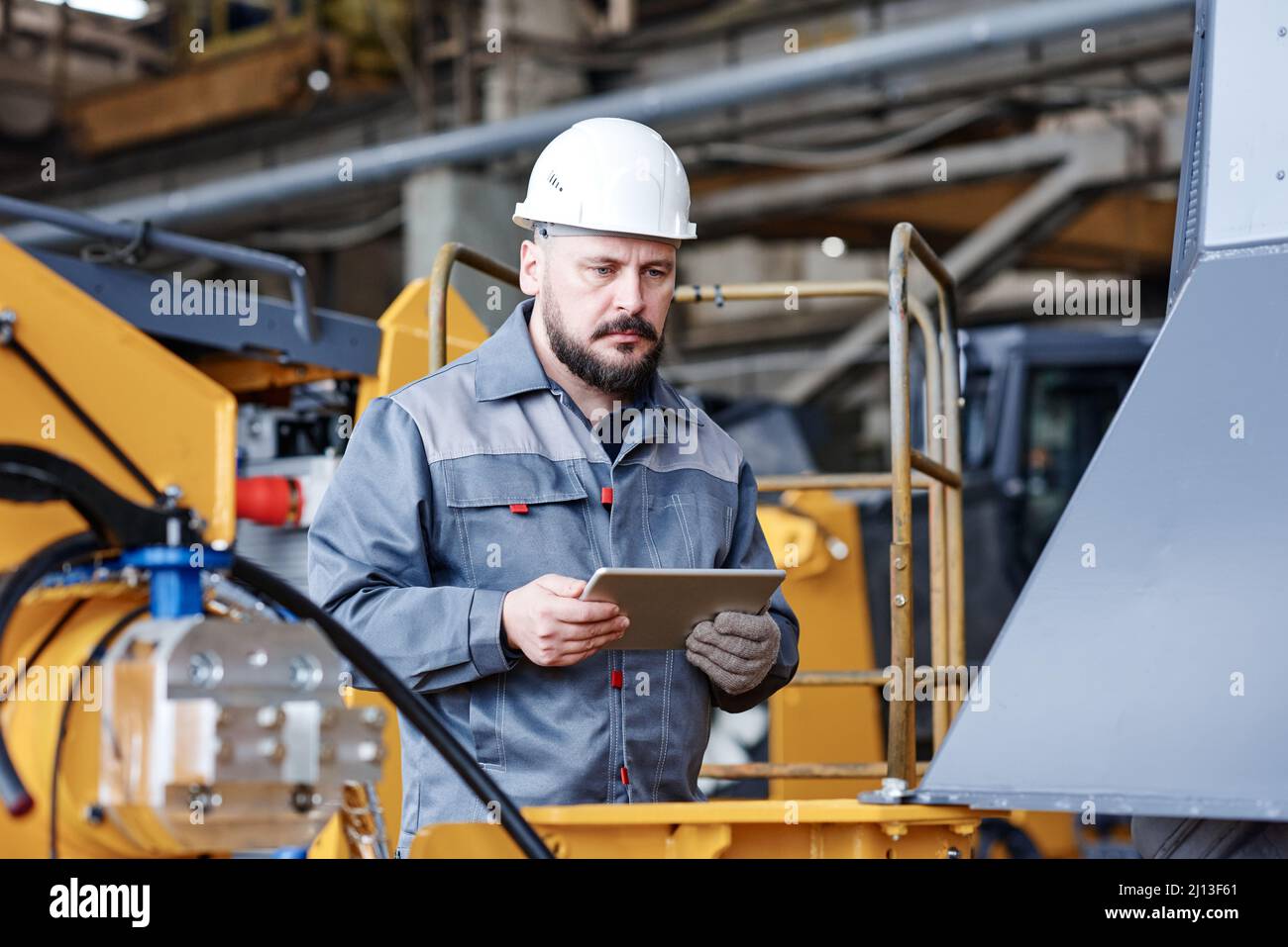 Mature serious engineer or quality control manager of industrial plant with tablet standing in ...