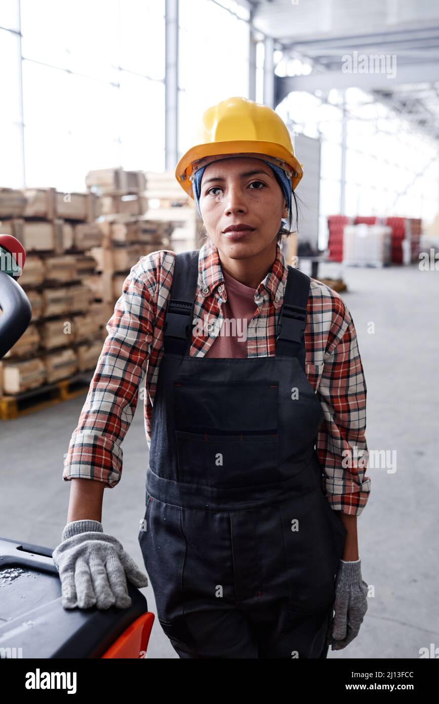 Young serious female engineer in workwear standing by cart with package ...