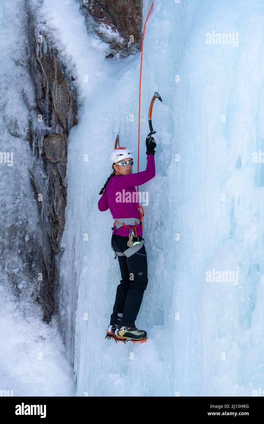 A female ice climber ascending an ice wall using ice axes and crampons ...