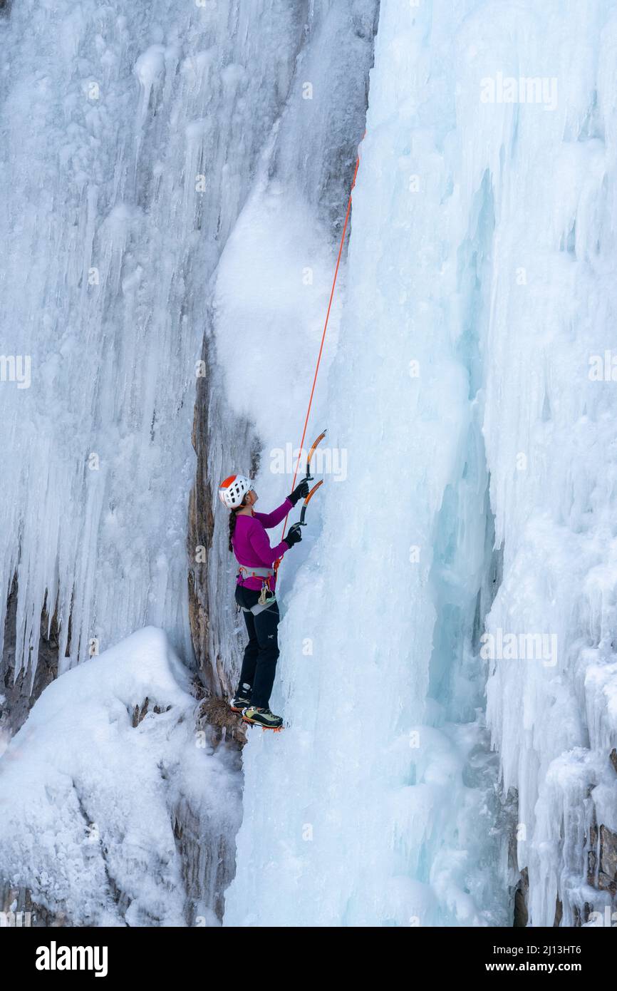 A female ice climber ascending an ice wall using ice axes and crampons ...
