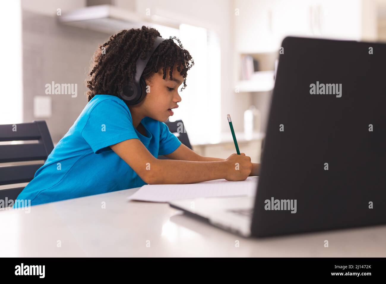 Hispanic boy wearing headphones writing in book sitting with laptop at ...