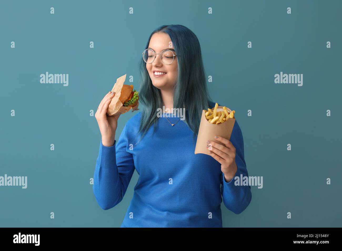 Young woman with unusual hair holding burger and french fries on color ...