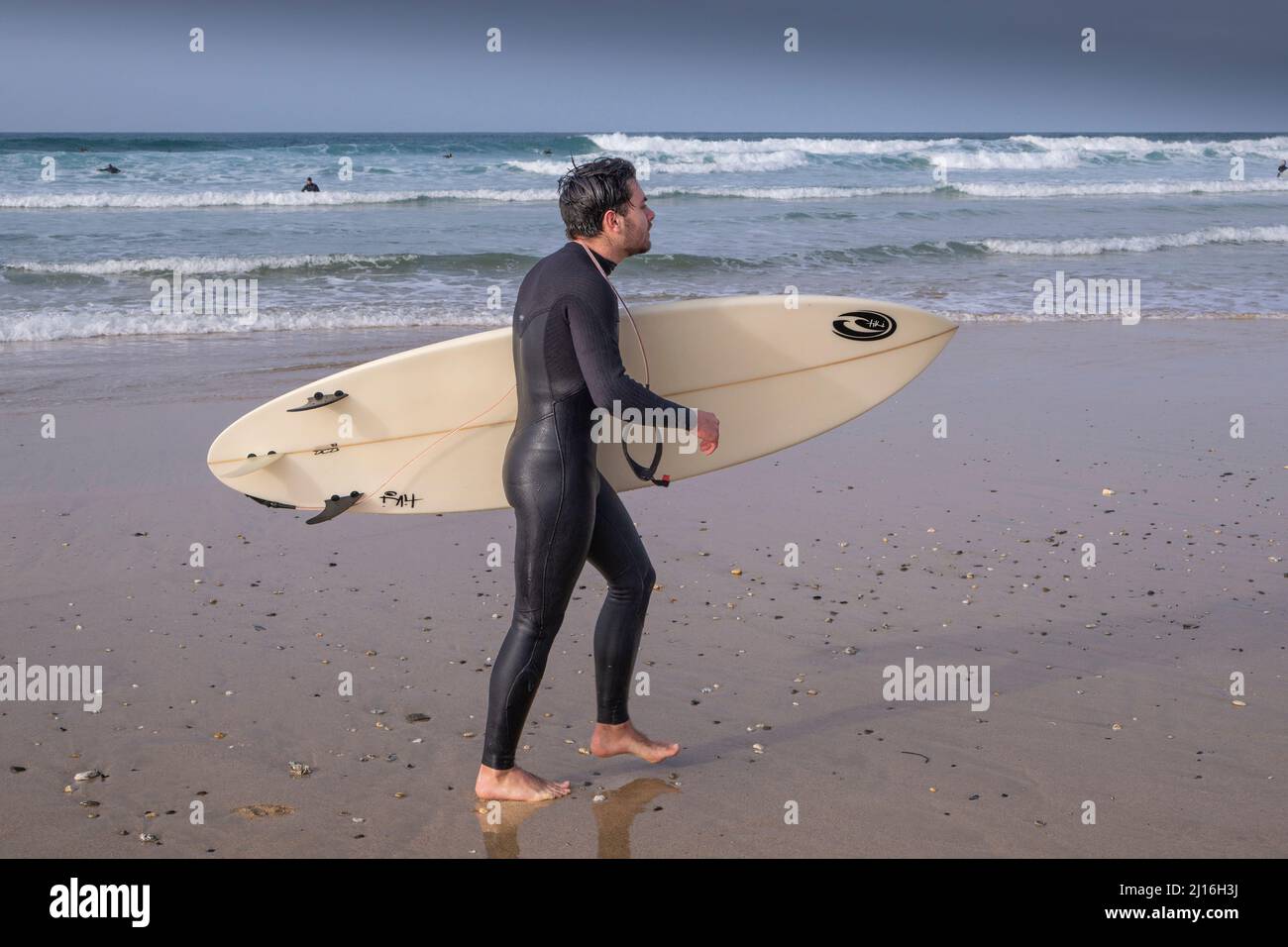 A surfer carrying his surfboard along the shoreline of Fistral Beach in ...