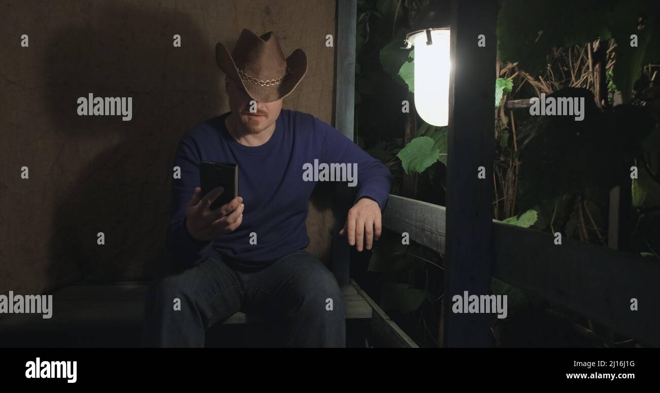 Cowboy man communicates online using his phone while sitting on porch ...