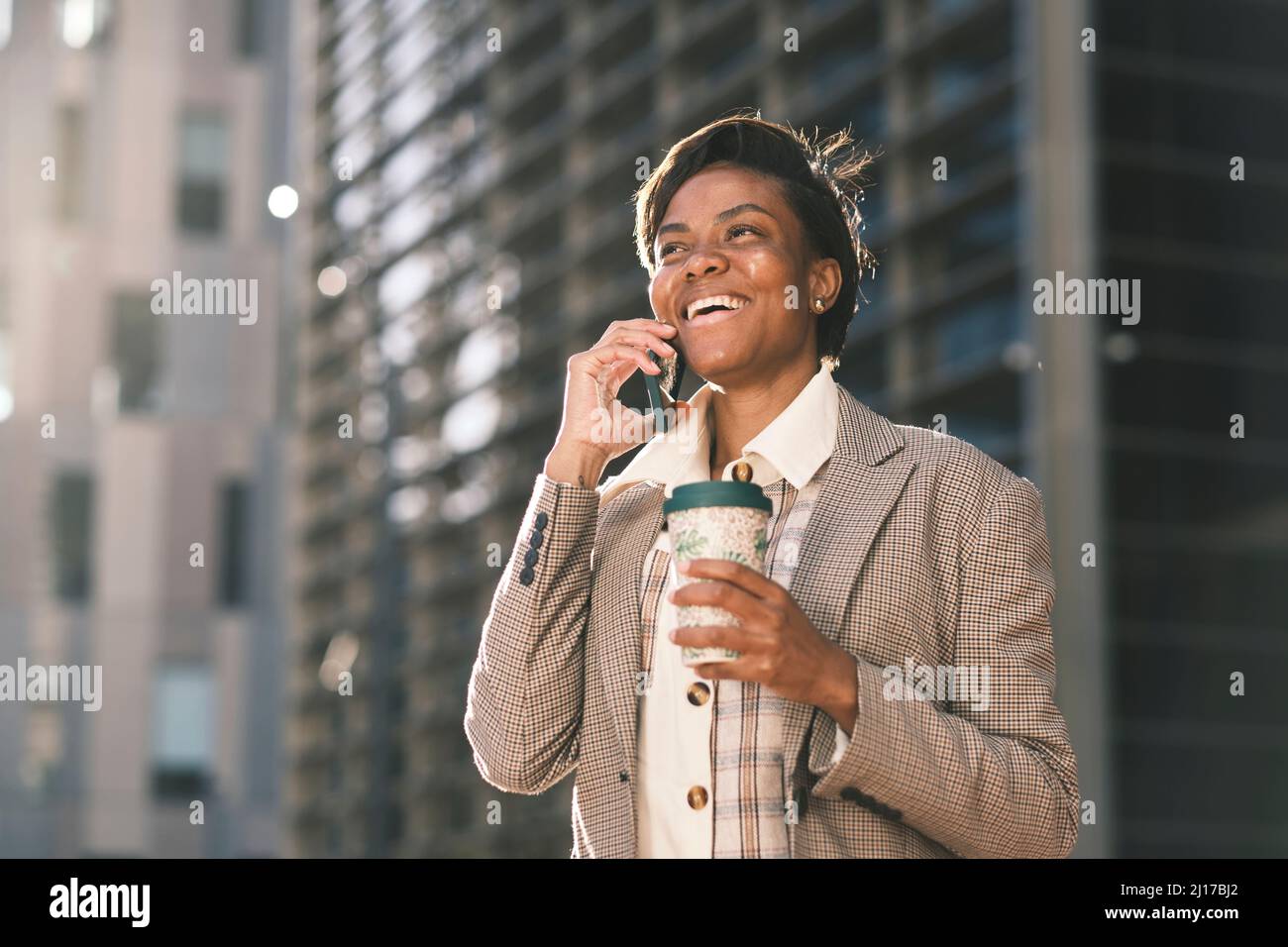 Cheerful businesswoman holding coffee cup talking through mobile phone ...