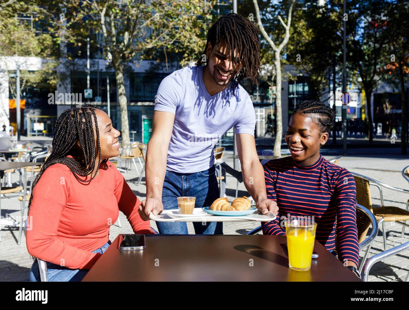 Happy young man carrying tray on table for friends at sidewalk cafe ...