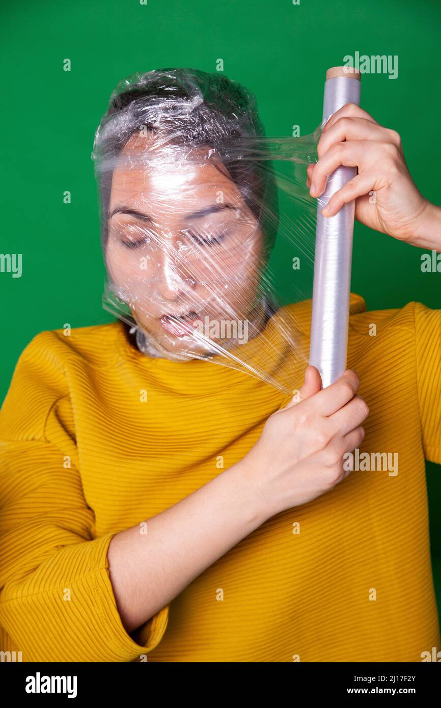 Woman wrapping plastic foil around face against green background Stock ...