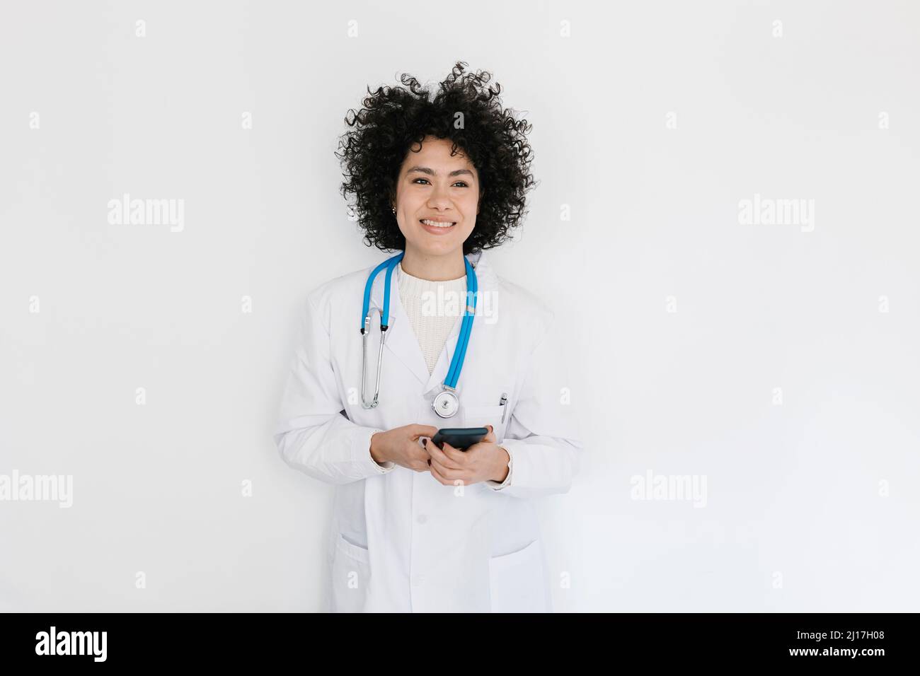 Smiling doctor holding mobile phone standing in front of white wall ...