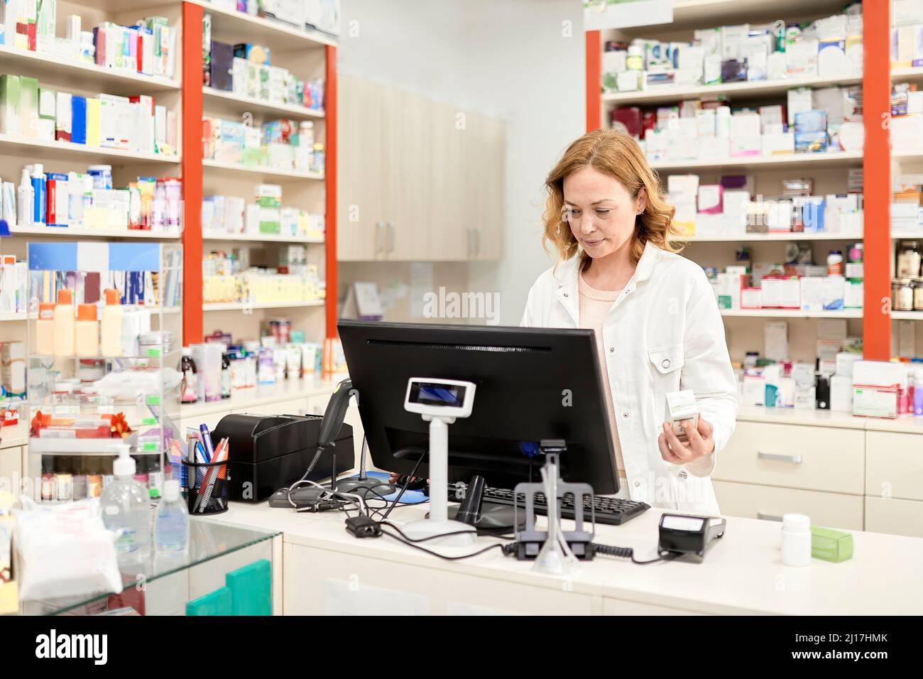Female pharmacist holding box of medicine using computer at checkout ...