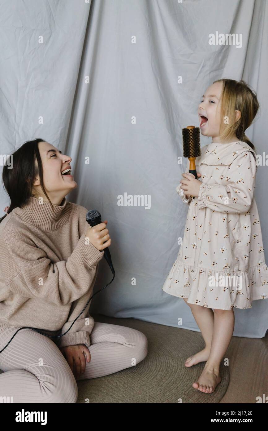 Cheerful mother with microphone looking at daughter holding hairbrush ...
