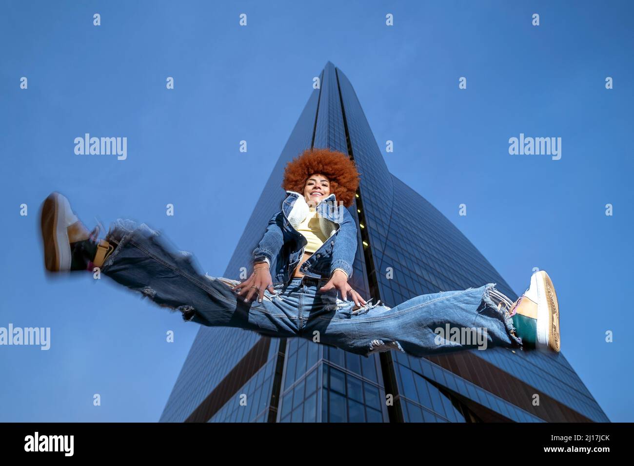 Smiling woman in redhead Afro hairstyle jumping in front of building at ...