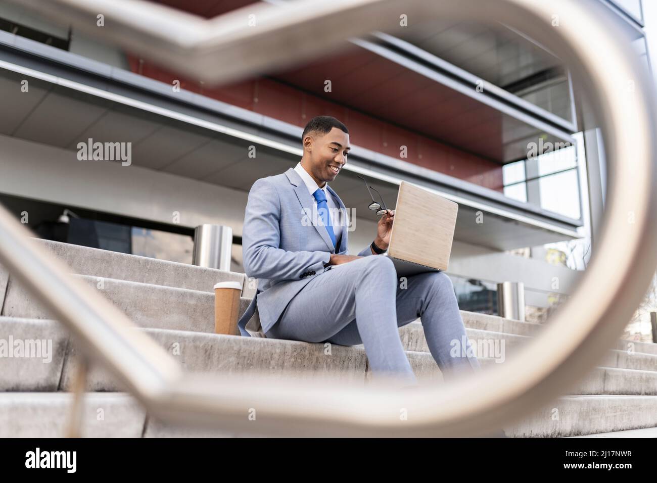 Smiling businessman using laptop sitting on steps in front of building ...