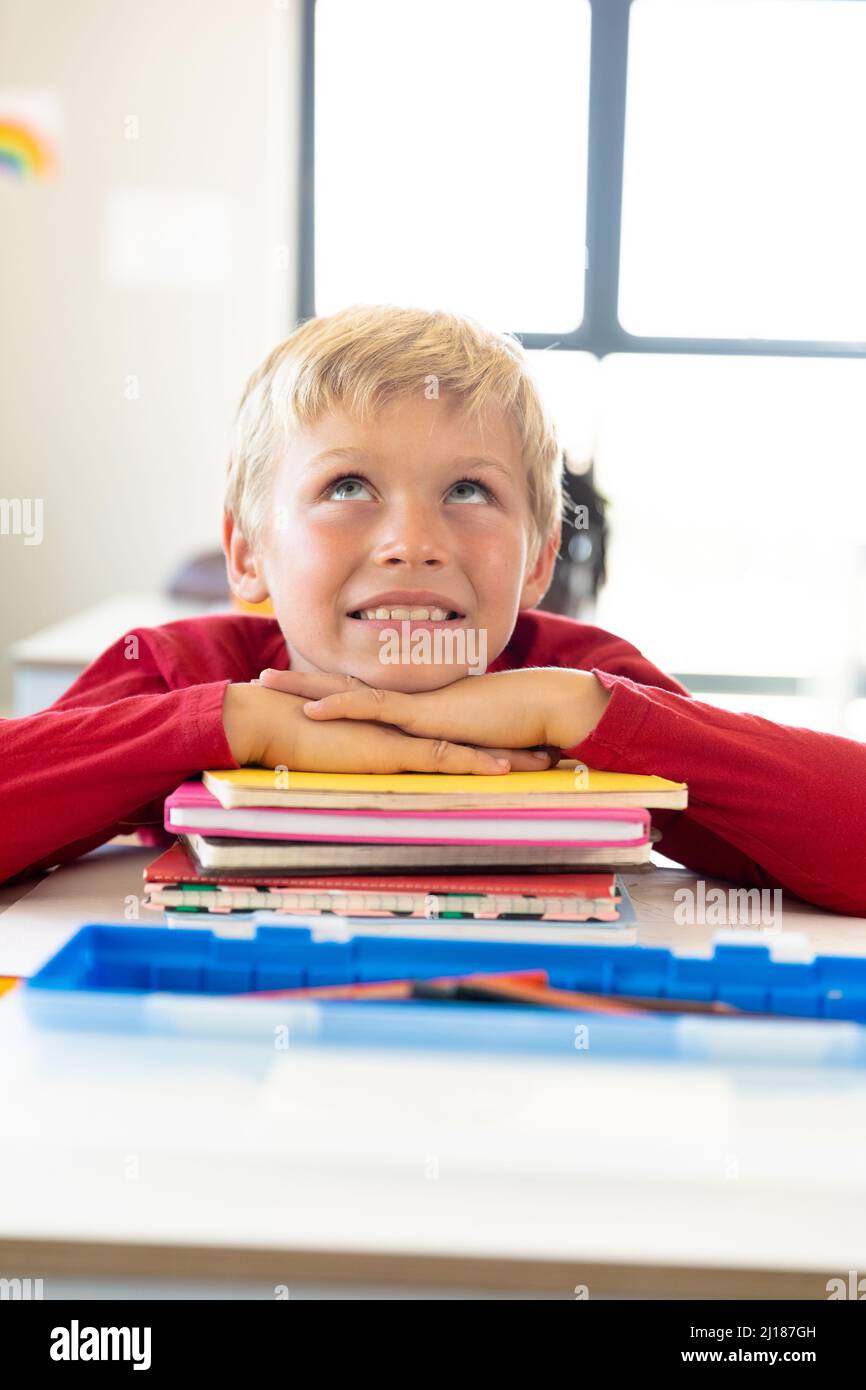Thoughtful caucasian elementary schoolboy leaning on stacked books at ...
