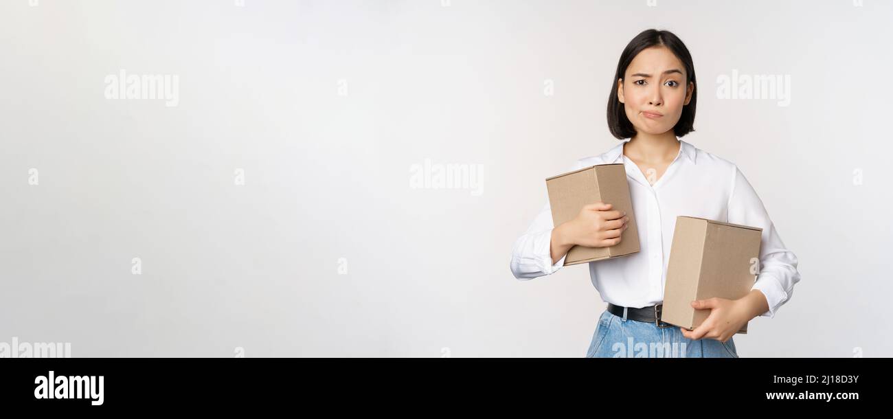 Complicated young asian woman holding two boxes, looking doubtful at ...