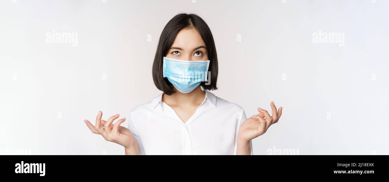 Annoyed asian woman in medical face mask, shrugging and looking up with ...