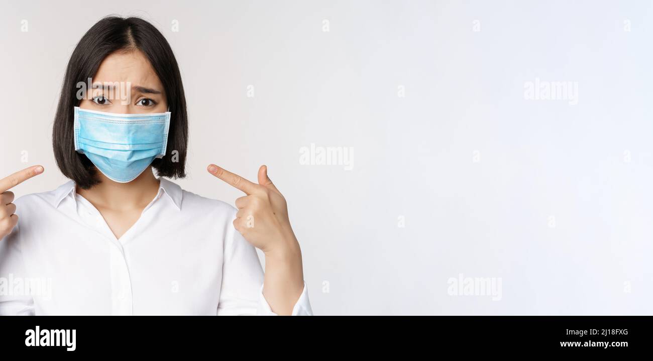 Close up of sad asian girl in medical face mask pointing at her head ...