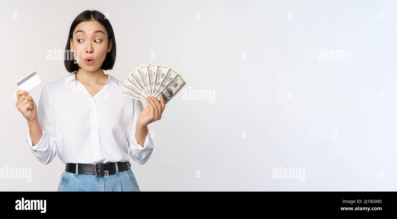 Excited korean girl looking at credit card, holding money cash, posing ...