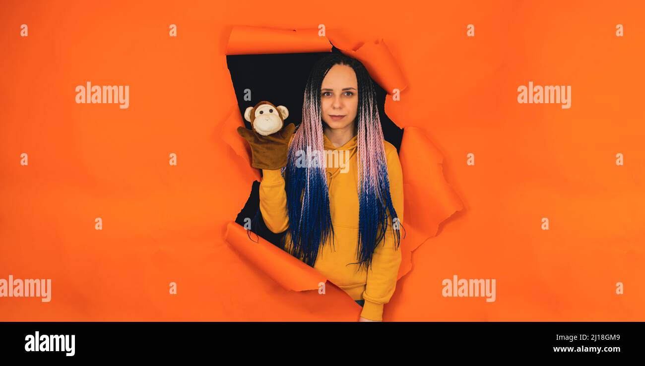 Young woman with soft puppet toy on hand looking out of hole of orange ...