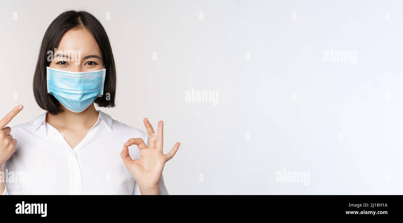 Portrait of asian girl in medical mask showing okay sign and pointing ...