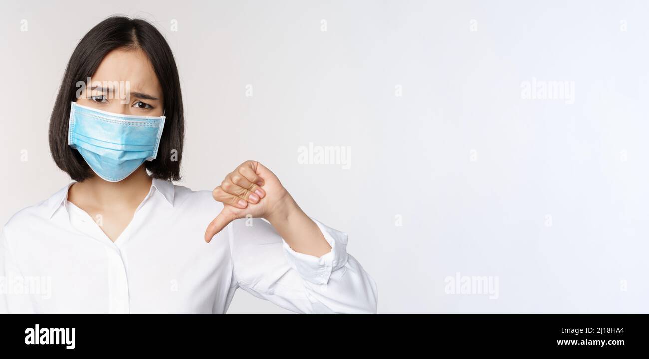 Portrait of asian woman in medical face mask showing thumbs down with ...
