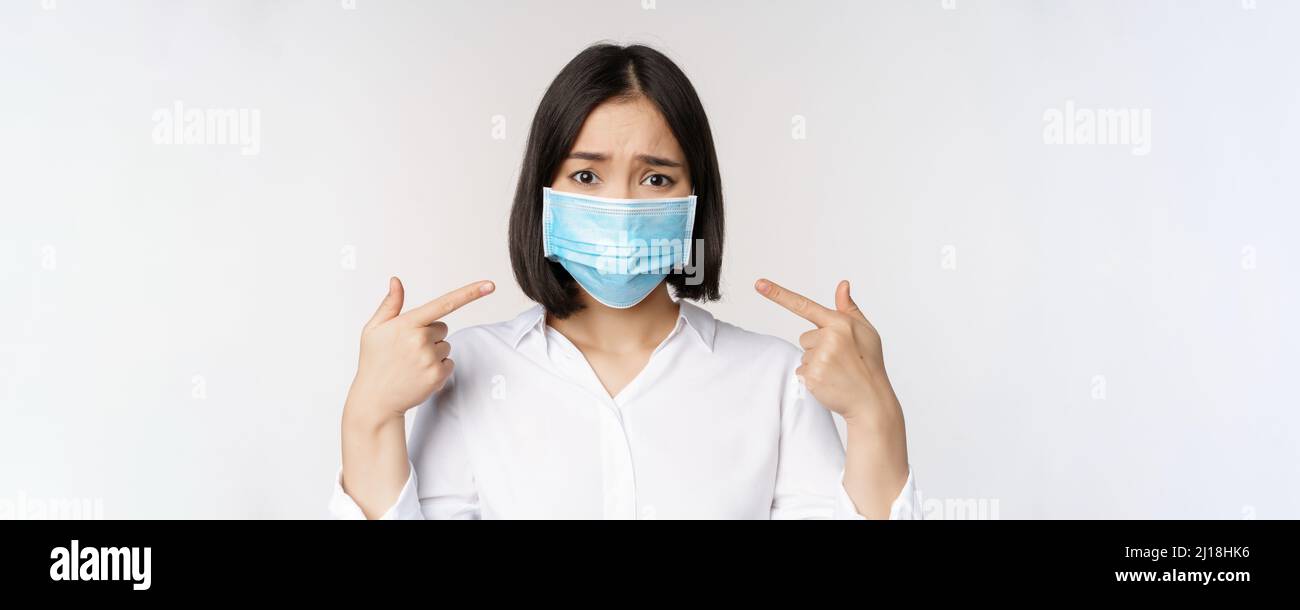 Close up of sad asian girl in medical face mask pointing at her head ...