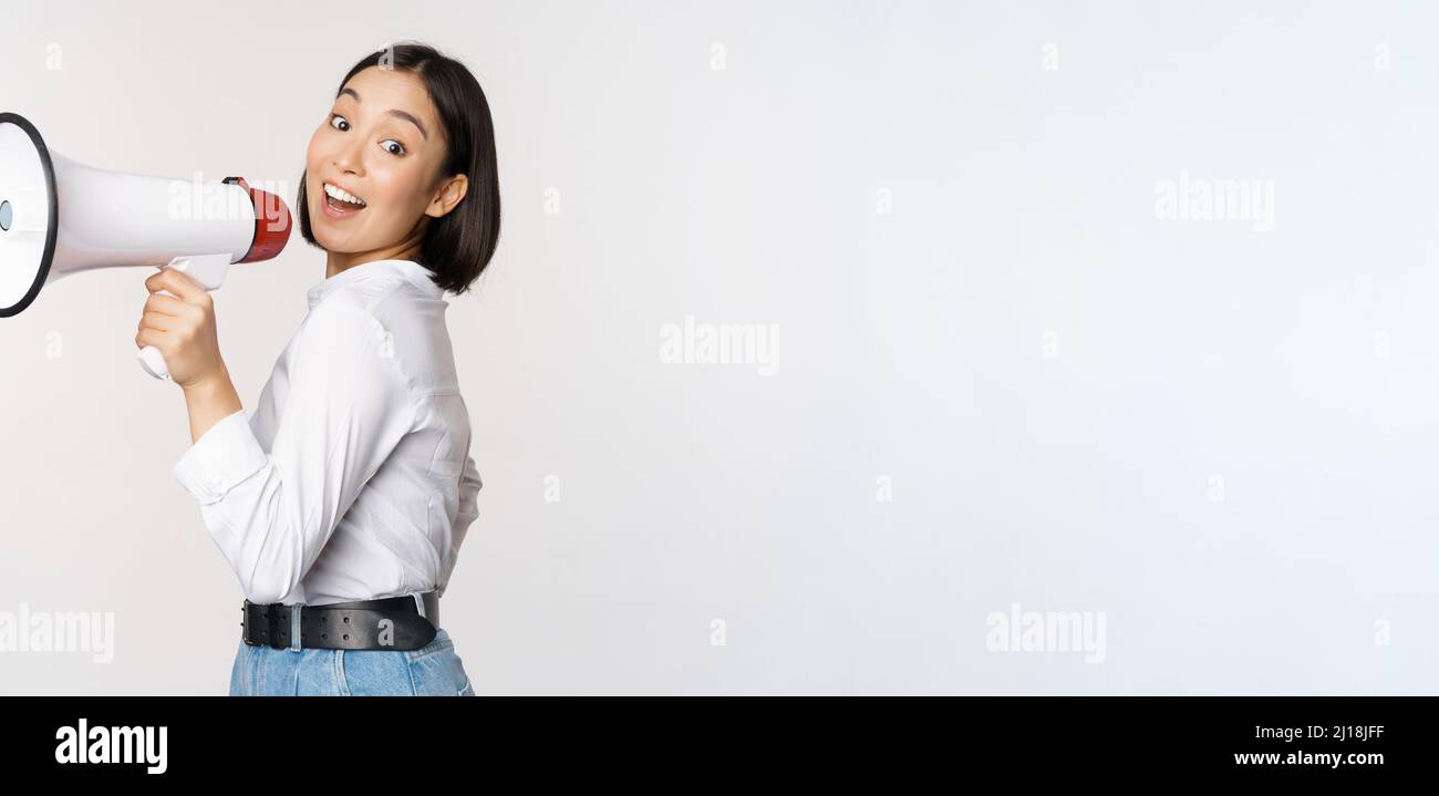 Beautiful young asian woman talking in megaphone, screams in speakerphone and smiling, making ...