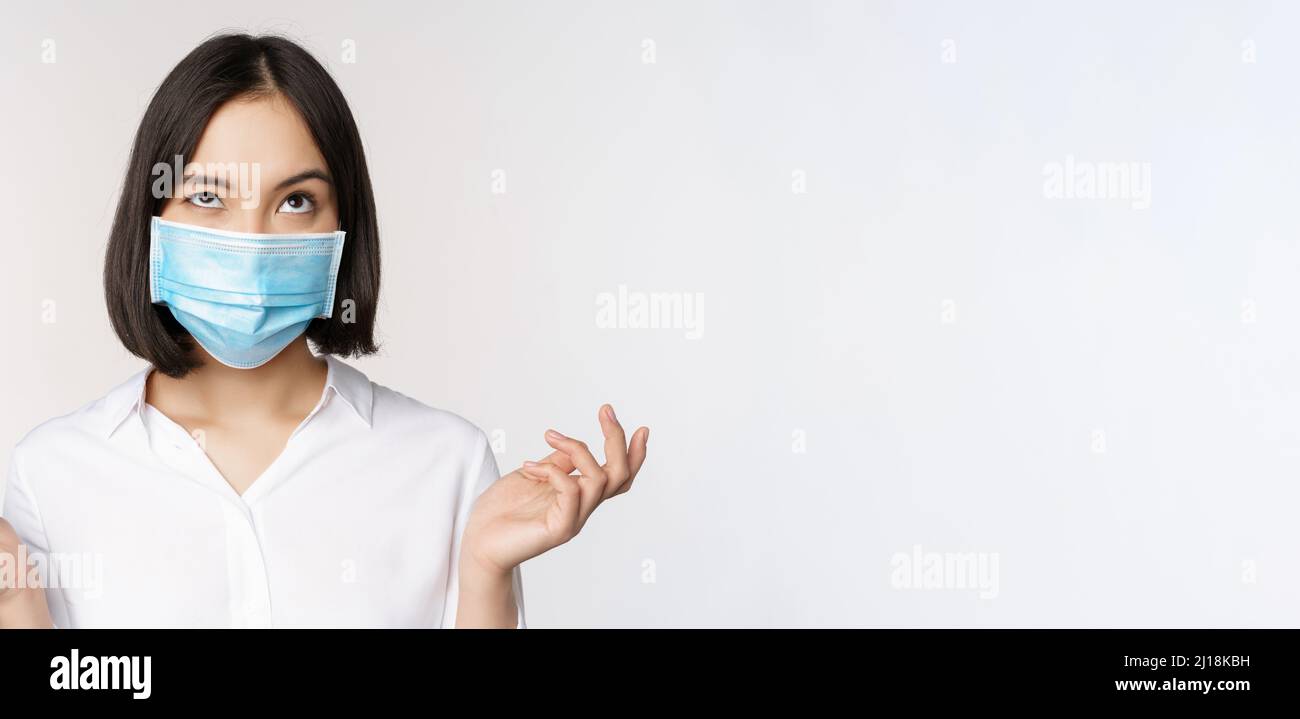 Annoyed asian woman in medical face mask, shrugging and looking up with ...