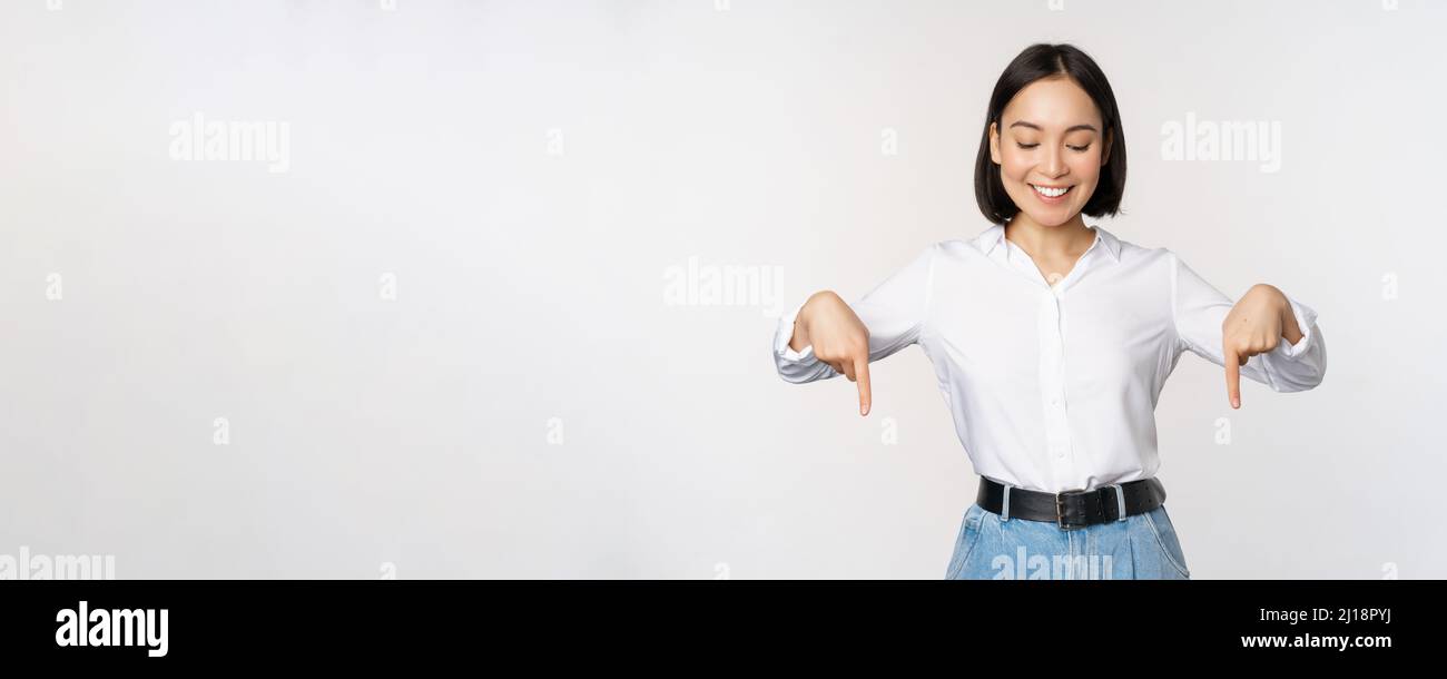 Portrait of happy asian woman pointing fingers down and looking below ...