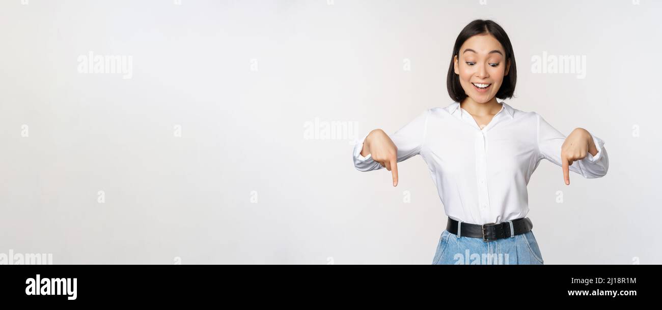 Portrait of happy asian woman pointing fingers down and looking below ...