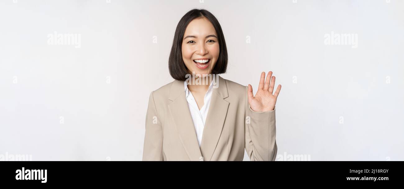 Friendly business woman, asian office lady waving hand and saying hello ...