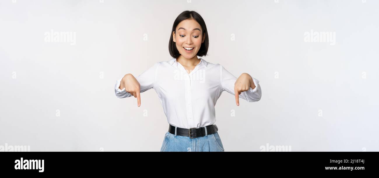 Portrait of happy asian woman pointing fingers down and looking below ...