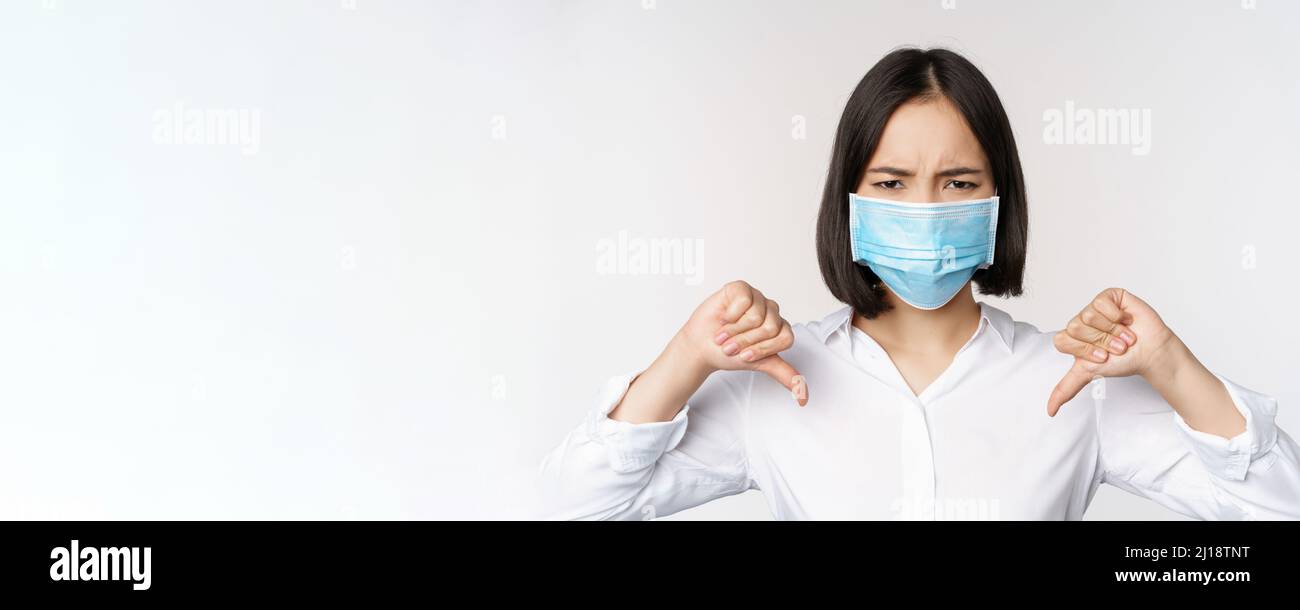 Portrait of asian woman in medical face mask showing thumbs down with ...