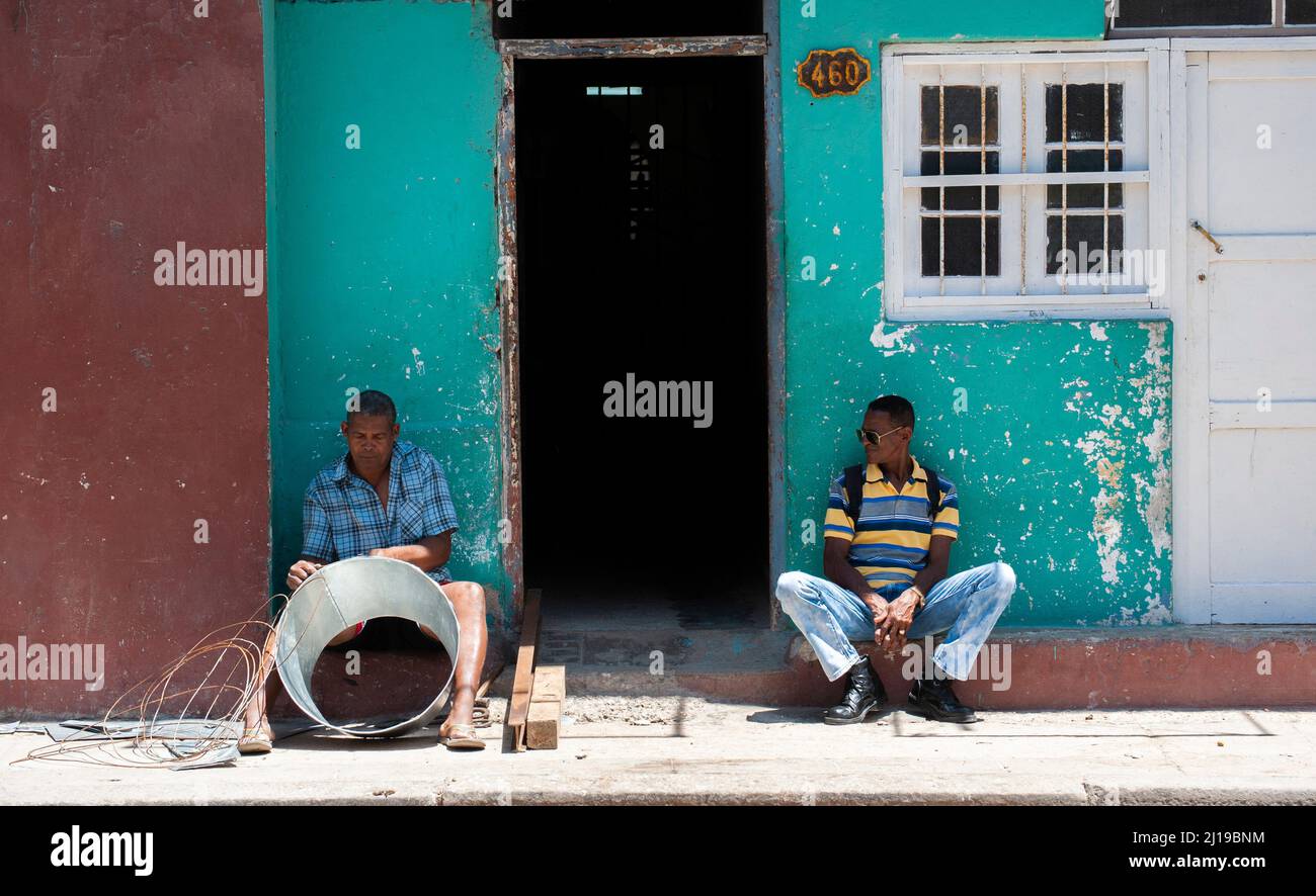Two men sit while one works making a type of metal tin with wires in ...