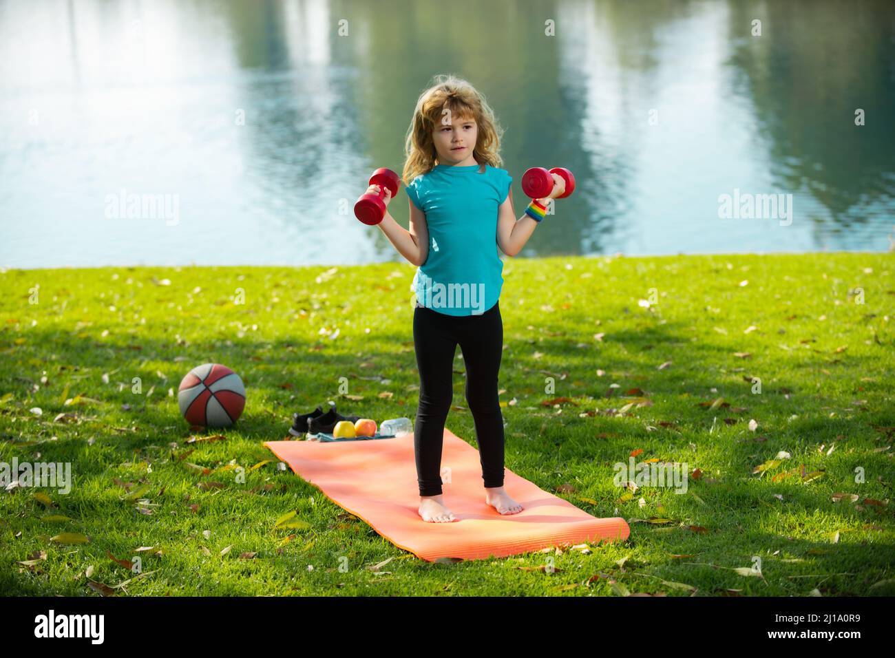 Cute child boy pumping up arm muscles with dumbbell outdoor in summer park. Fitness kids with ...